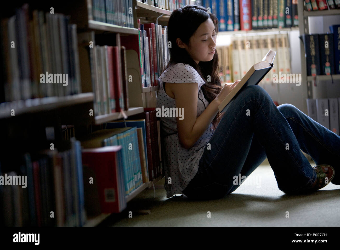 Young woman sitting on floor in library reading book Stock Photo - Alamy