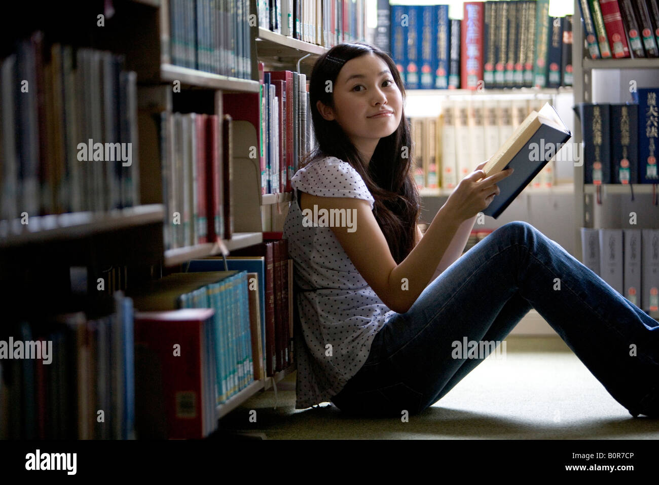 Young woman sitting on floor in library holding book Stock Photo - Alamy