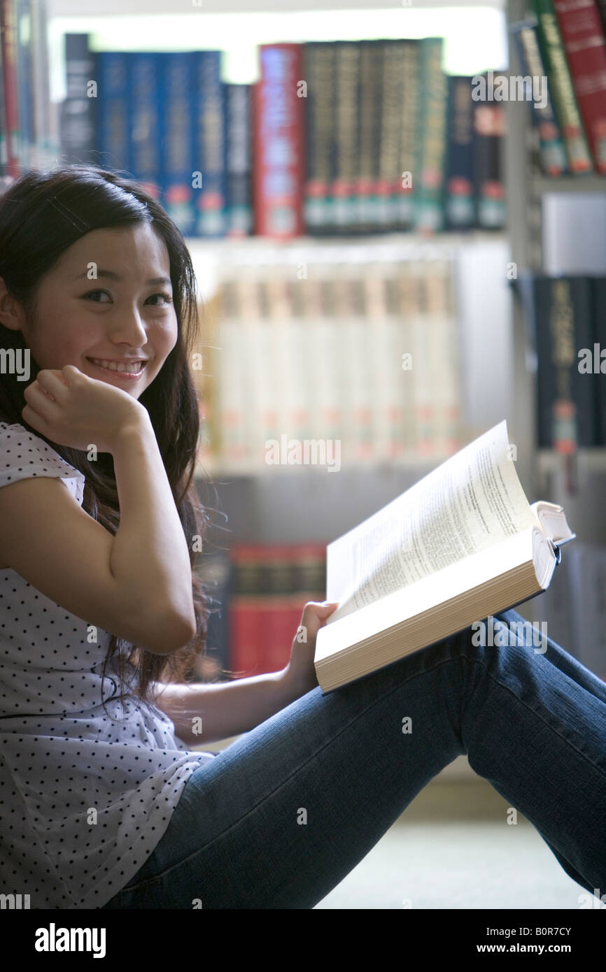 Portrait of young woman sitting on floor in library reading book Stock ...