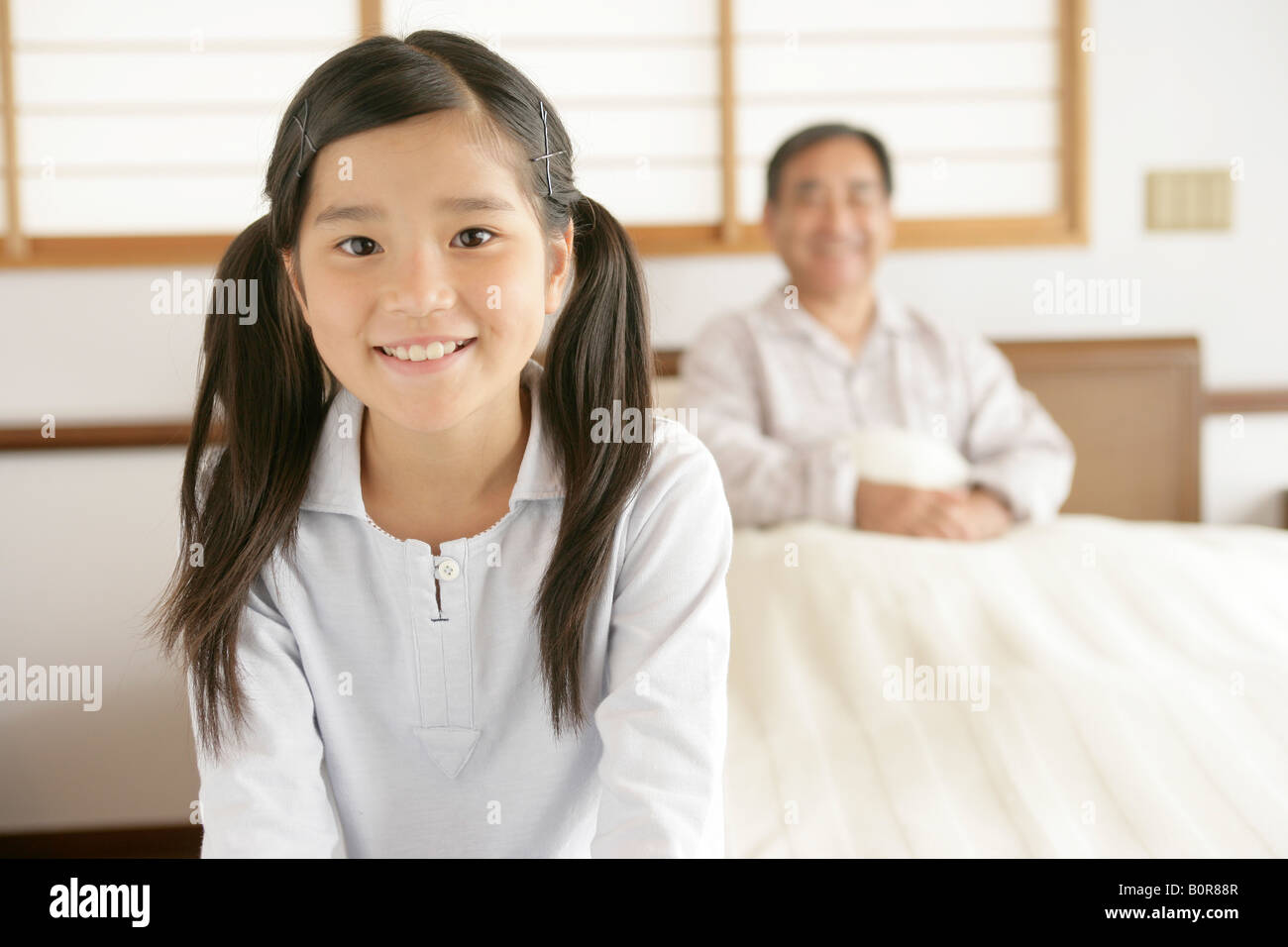 Portrait Of Girl Sitting At Bedside With Grandfather Sitting In Bed In Background In Nursing