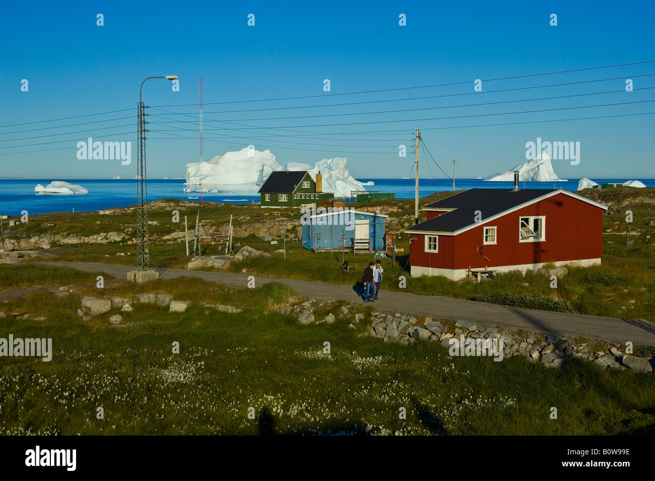 Inuit in front of a wooden house, icebergs visible at back ...