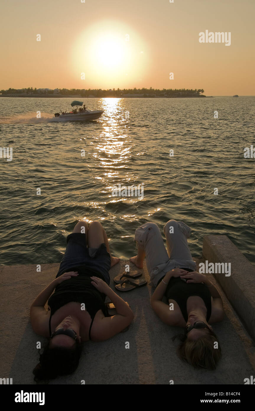 Two women friends recline on Mallory Square Pier as people wave from ...