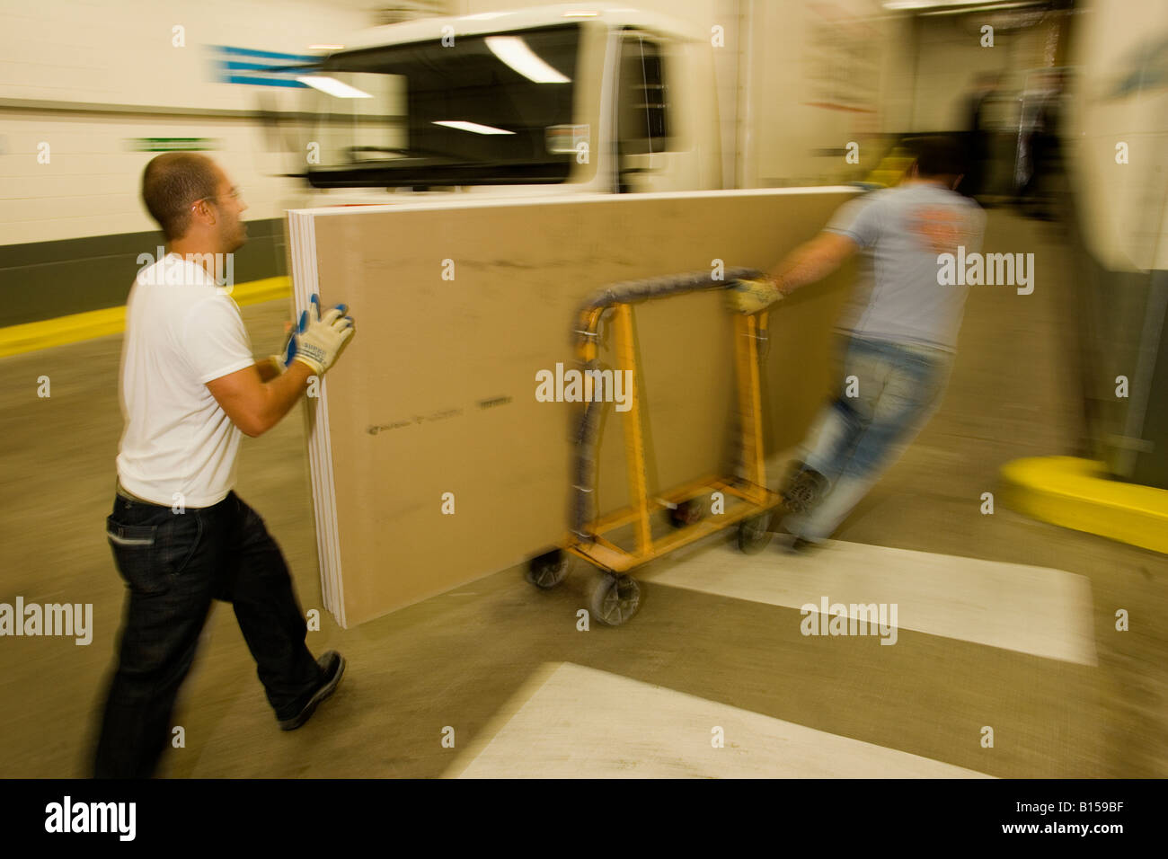 A pair of workmen move a large container on a small industrial cart ...