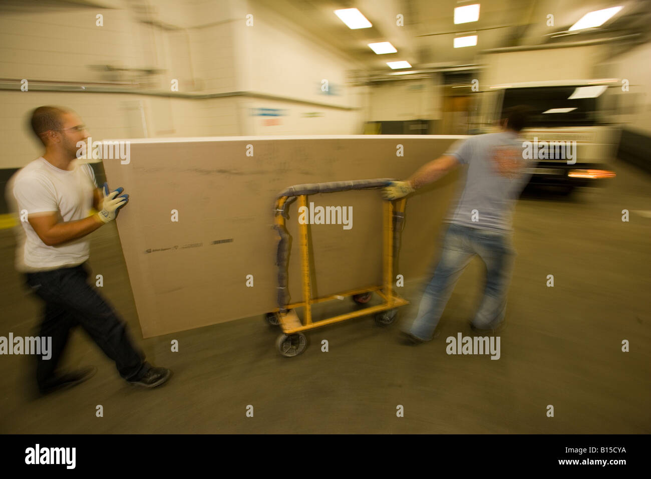 A pair of workmen move a large container on a small industrial cart ...