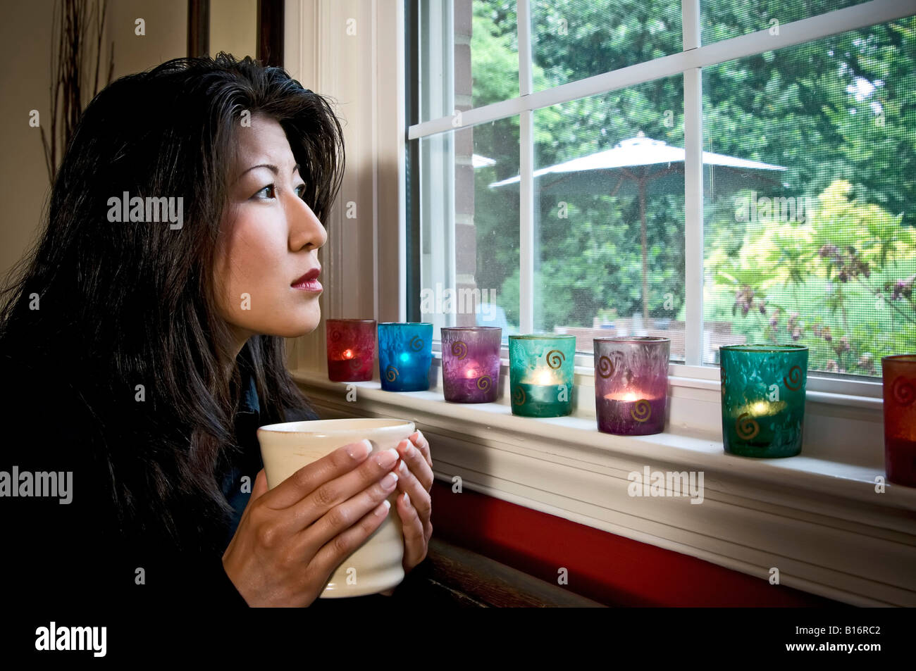 An Asian woman holds a large cup of tea while looking out a window ...