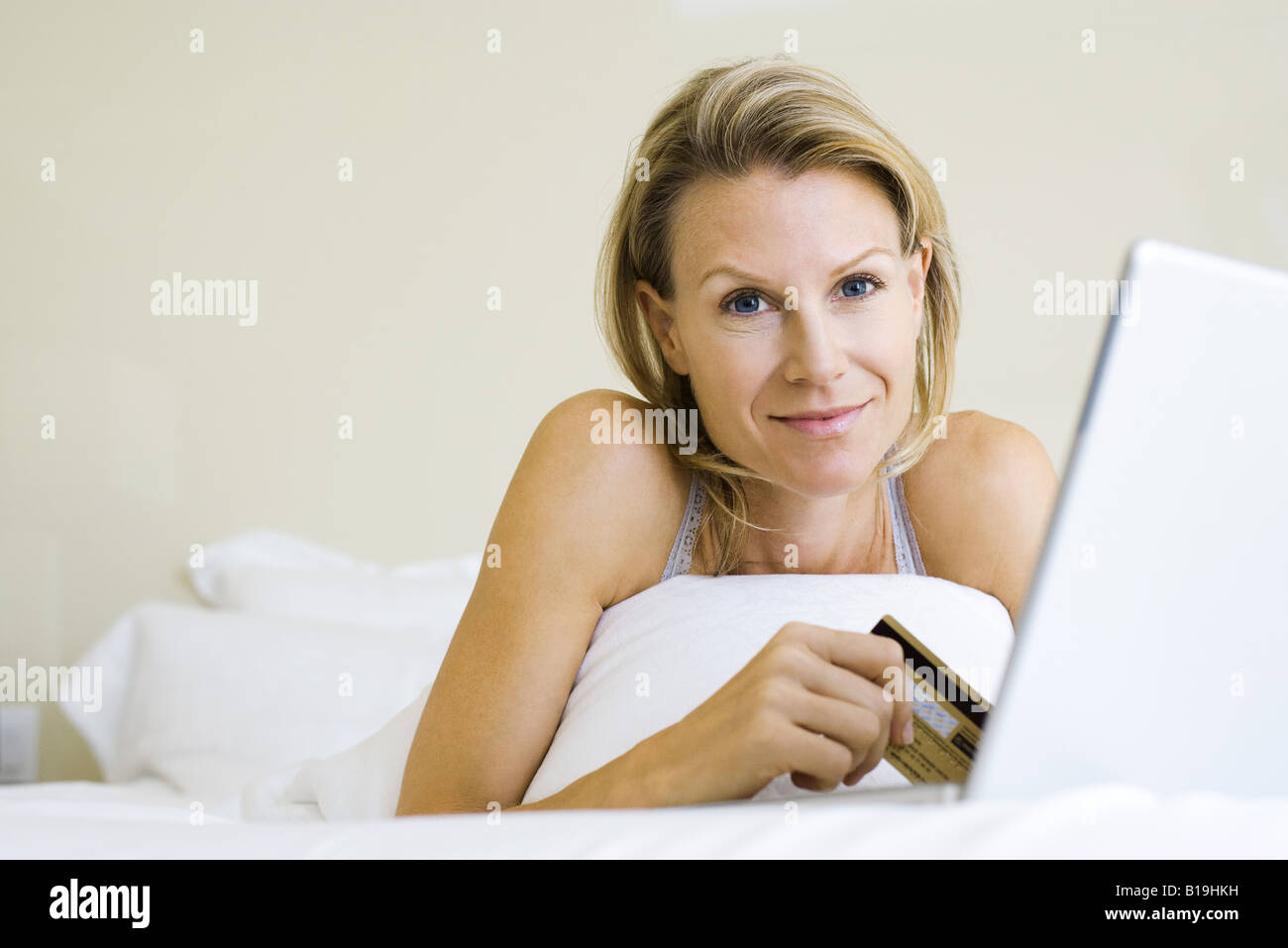 Woman lying on bed, using credit card and laptop, smiling at camera ...