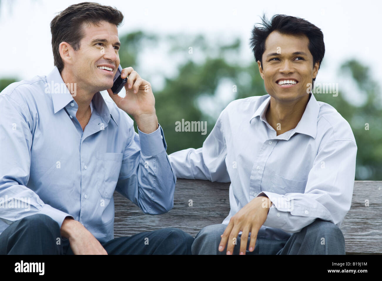 Two men sitting on bench, smiling, one using cell phone Stock Photo - Alamy