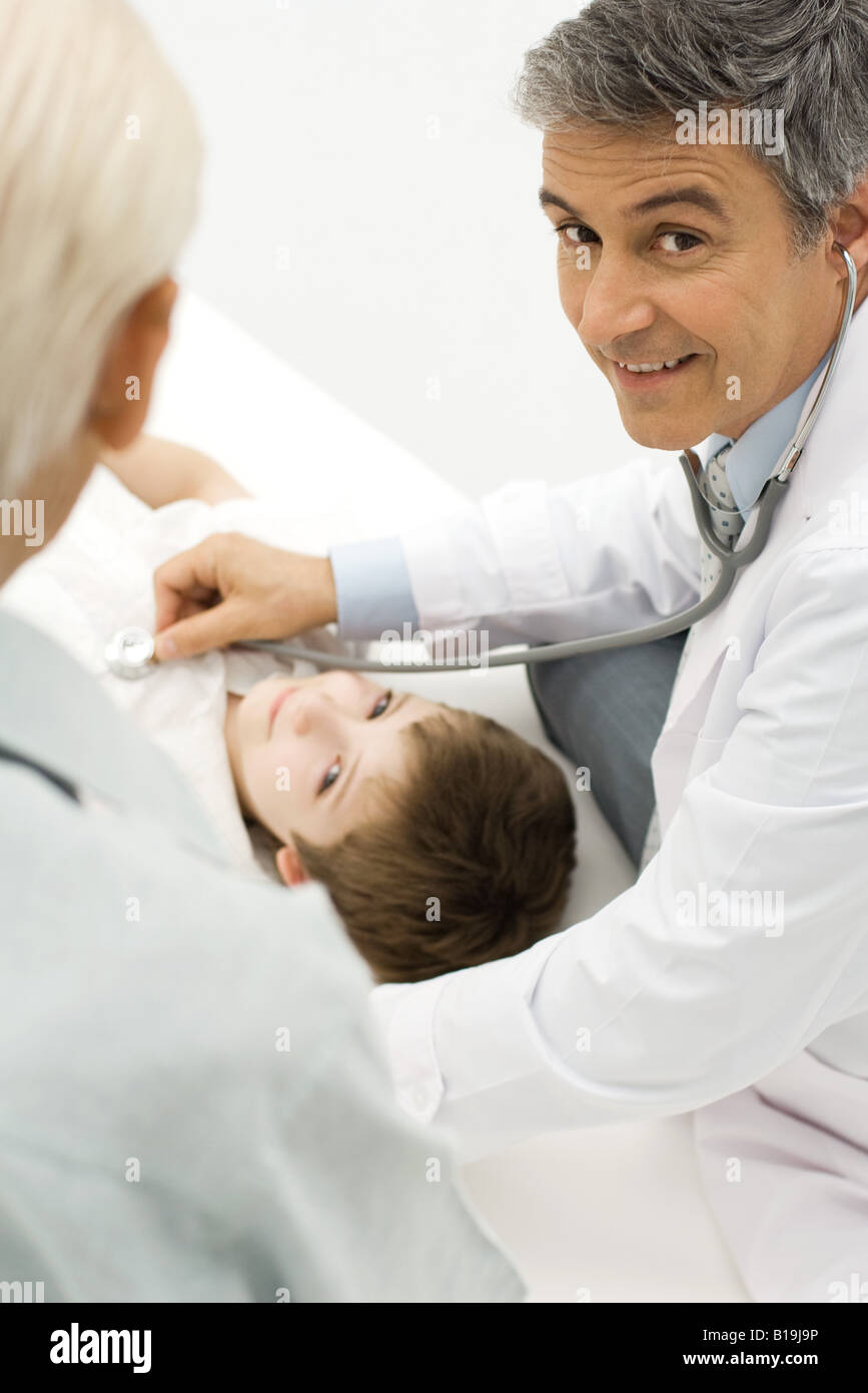 Doctor listening to young patient's heart, smiling over shoulder at ...