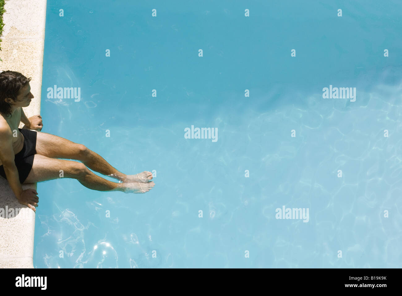 Man sitting on edge of swimming pool, soaking feet in water, high angle ...