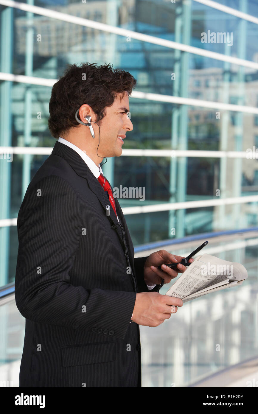 Businessman using mobile phone, reading newspaper outside office ...