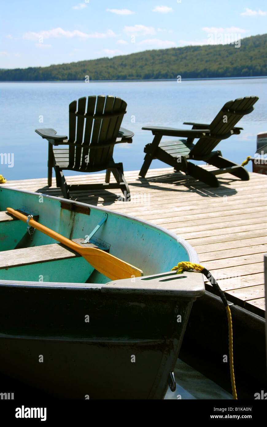 Paddle boat and two adirondack wooden chairs on dock facing a blue lake ...