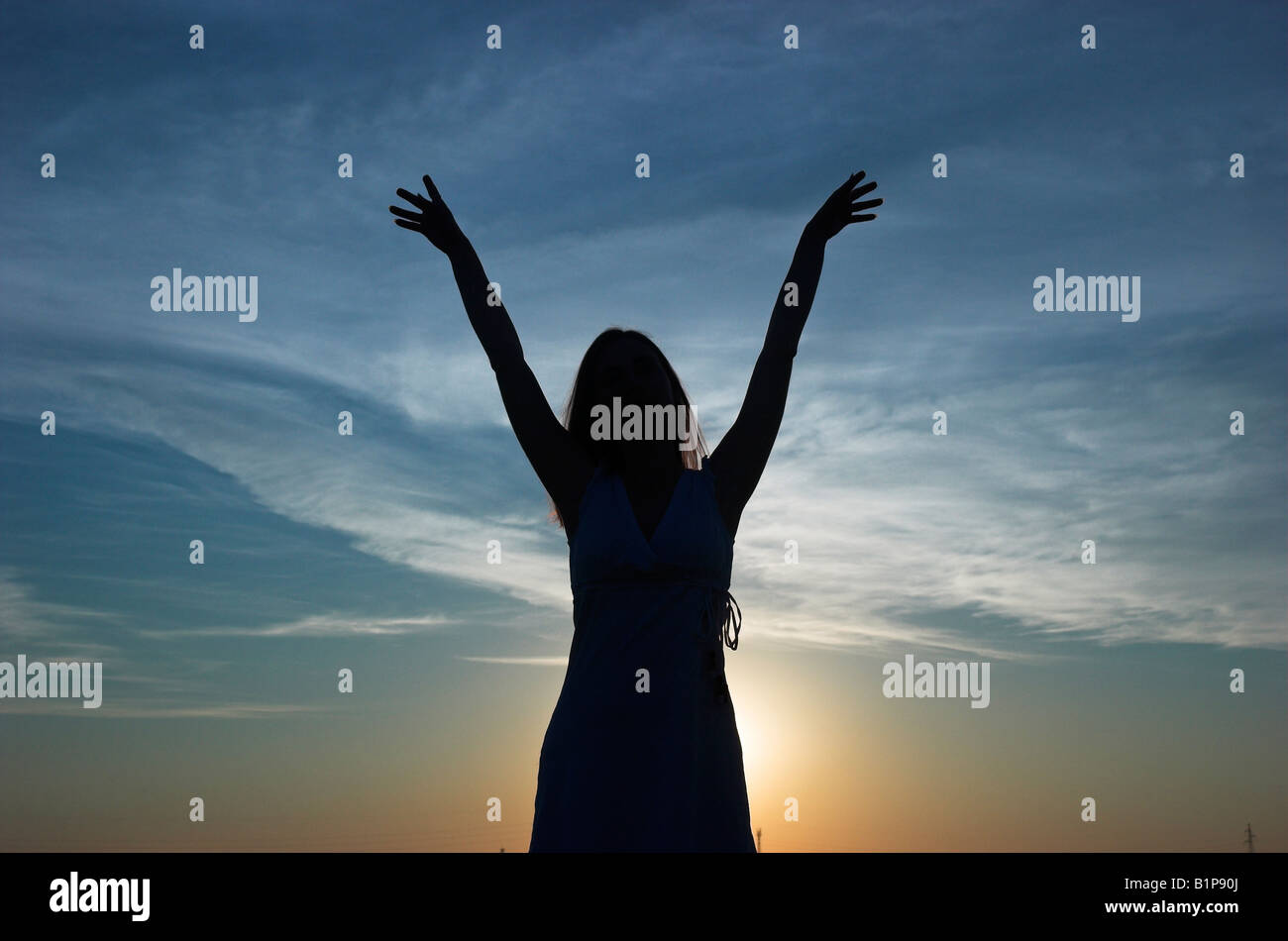 Silhouette of a young woman at dusk arms raised sun shining behind ...