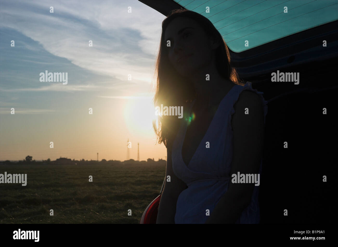 Silhouette of a young woman sitting on open car boot at dusk sun ...