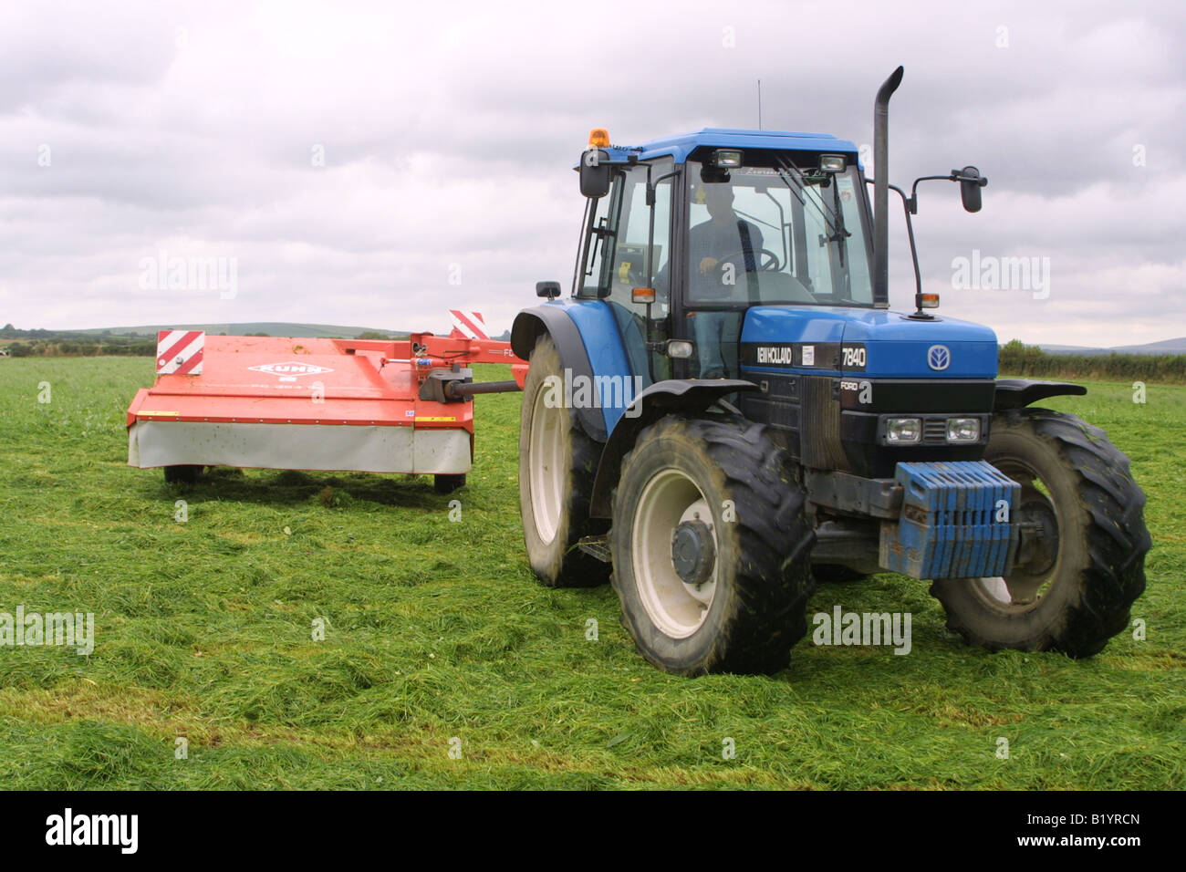 Blue Tractor and red farm machinery cutting grass for silage.Horizontal ...