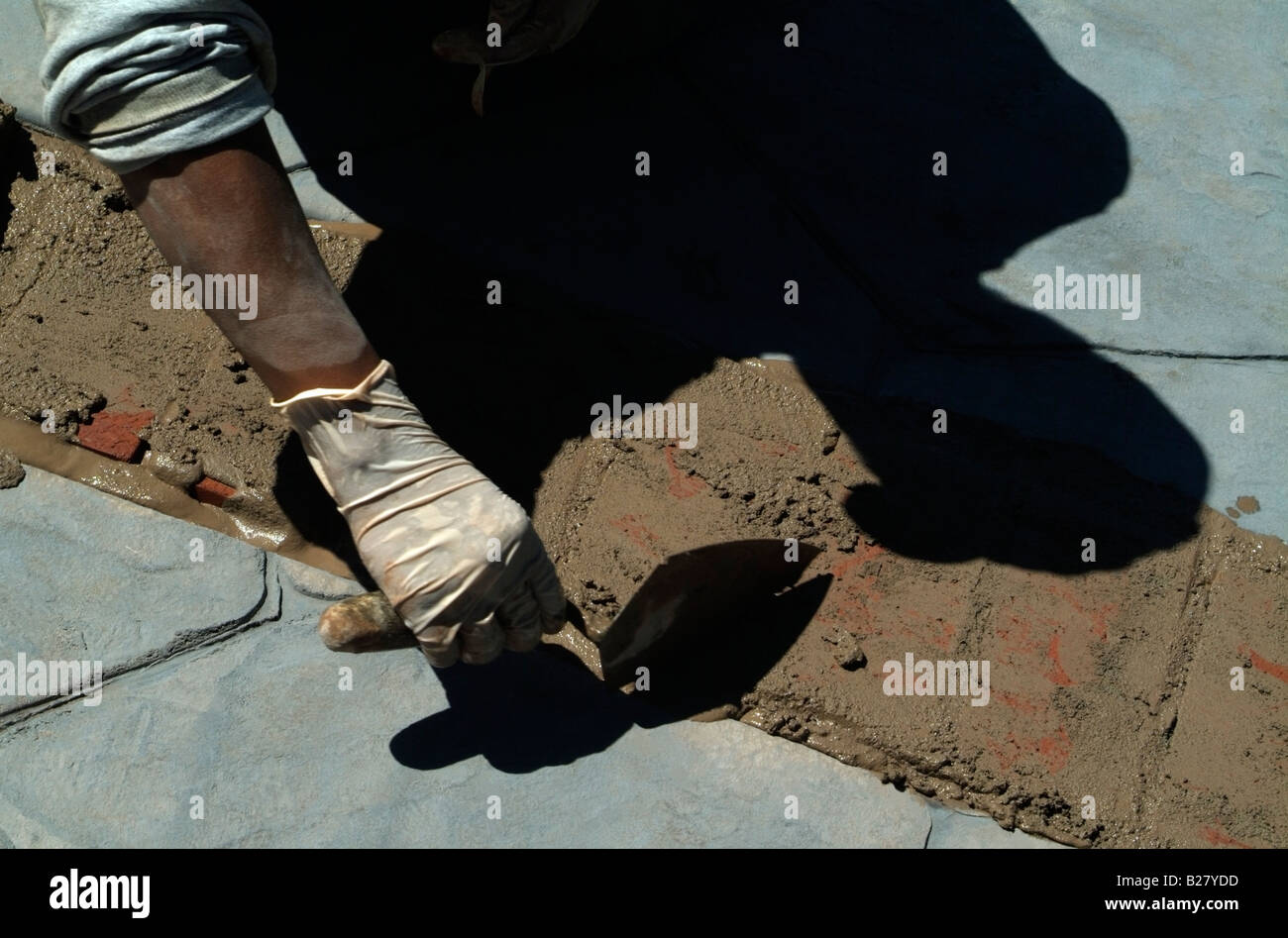 The gloved hands of a Mason using a trowel to place mortar between clay ...