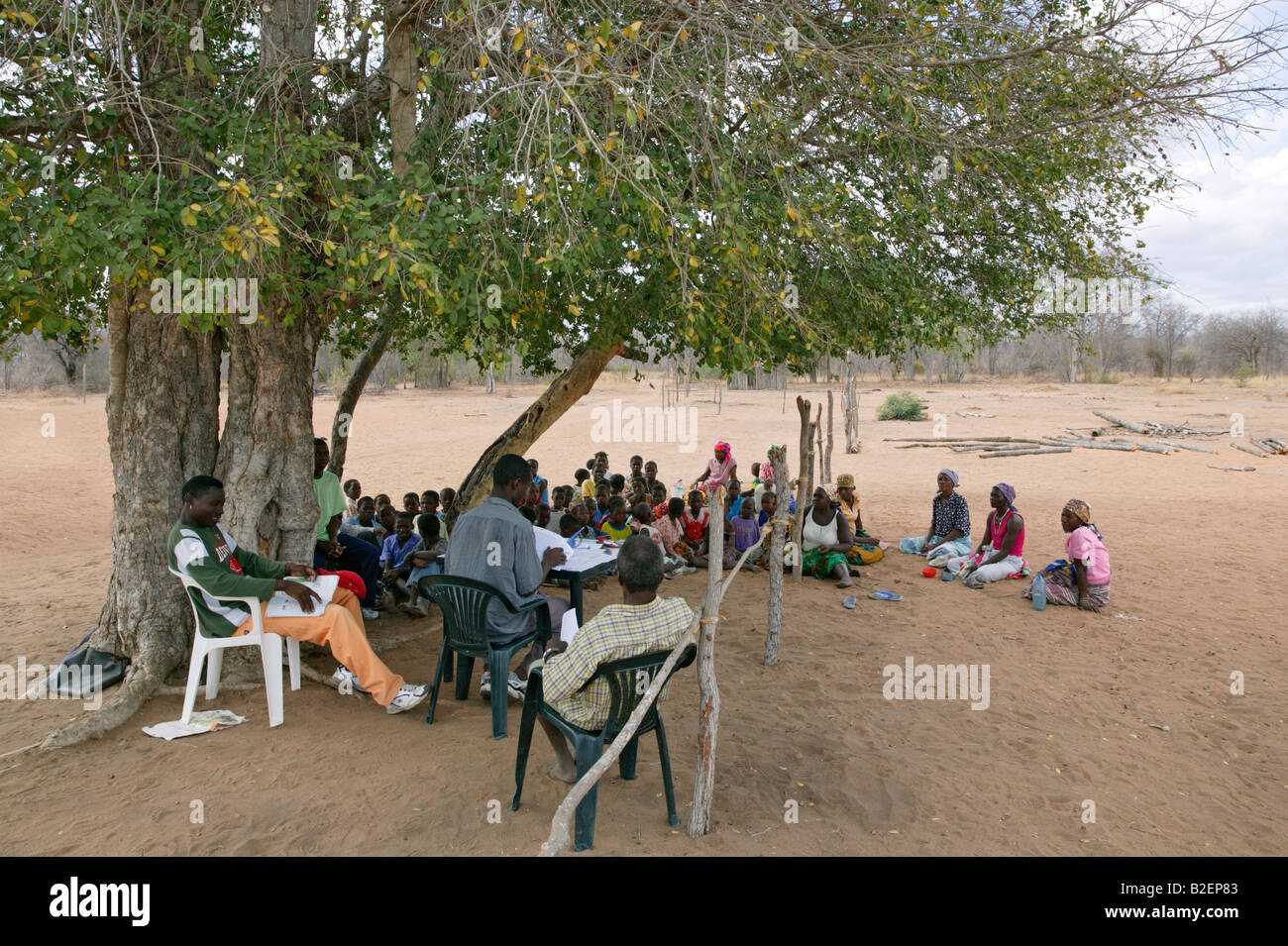 School children sitting in the sand under a tree while two school ...