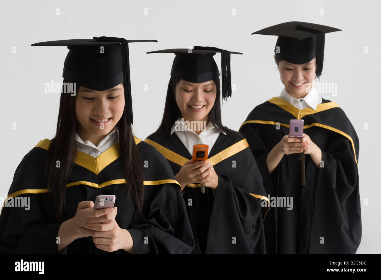 Three young female graduates text messaging with mobile phones and ...