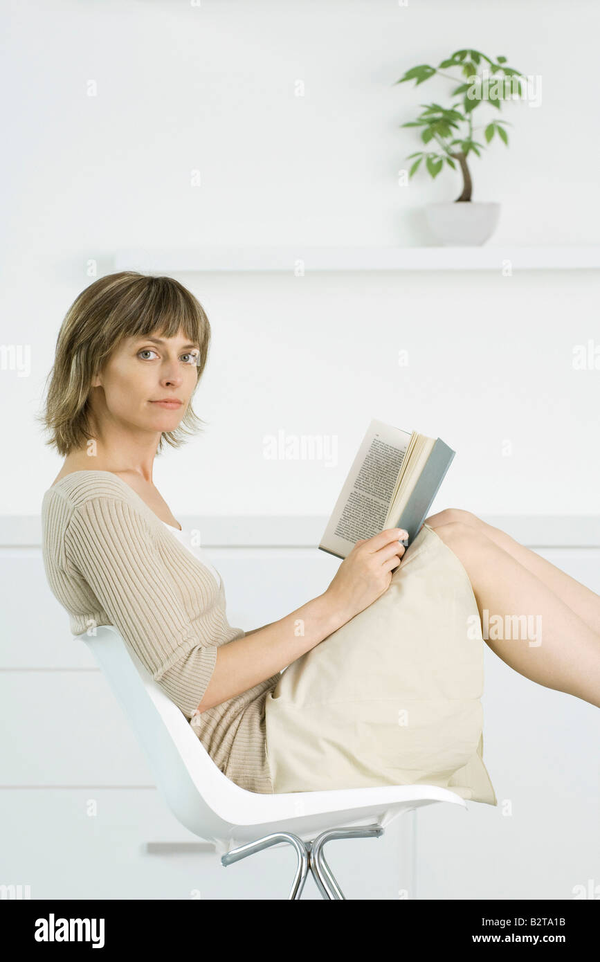 Woman leaning back in chair, holding book in her lap, looking at camera ...