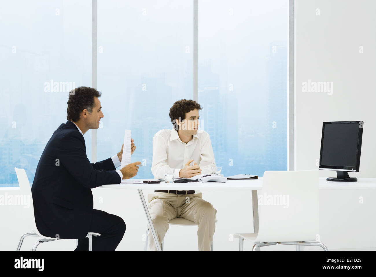 Businessmen sitting at table, looking at monitor, teleconferencing ...