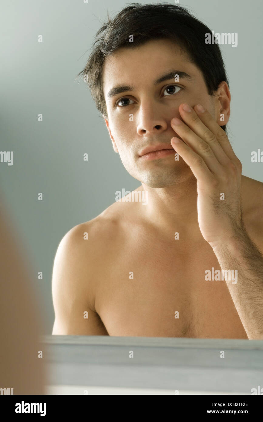 Bare-chested man looking at self in mirror, touching face Stock Photo ...