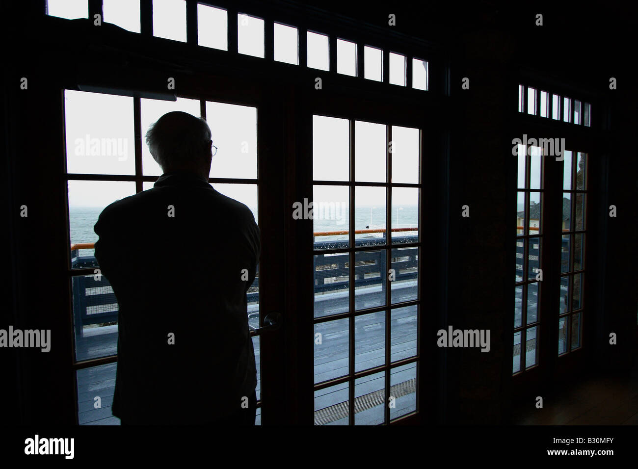 A man is cast in silhouette as he looks out balcony windows at the sea ...