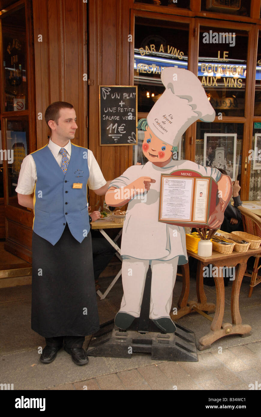 A garson (waiter) waiting for customers next to a cutout chef displayed ...