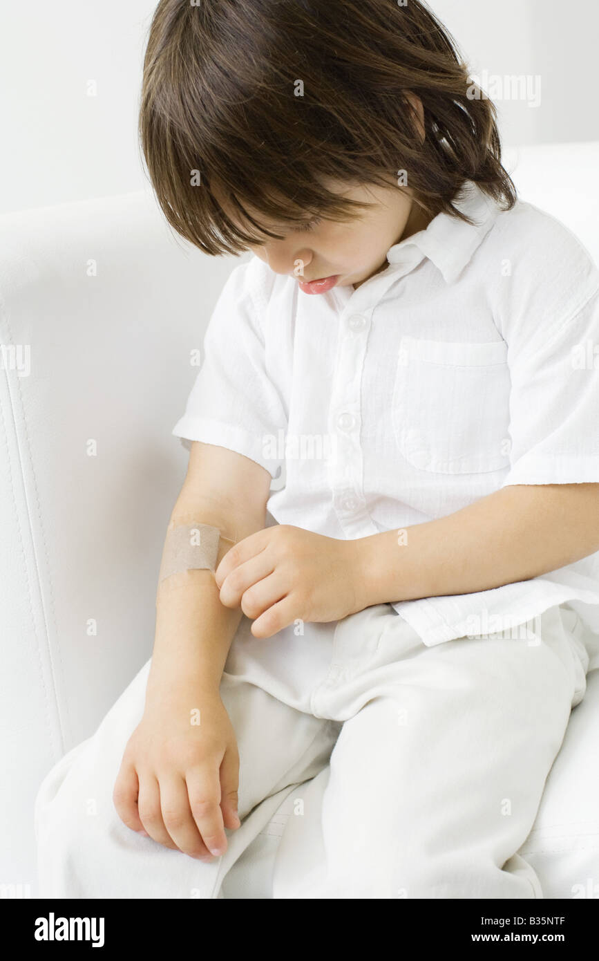 Little boy picking at adhesive bandage on his arm, head down Stock ...