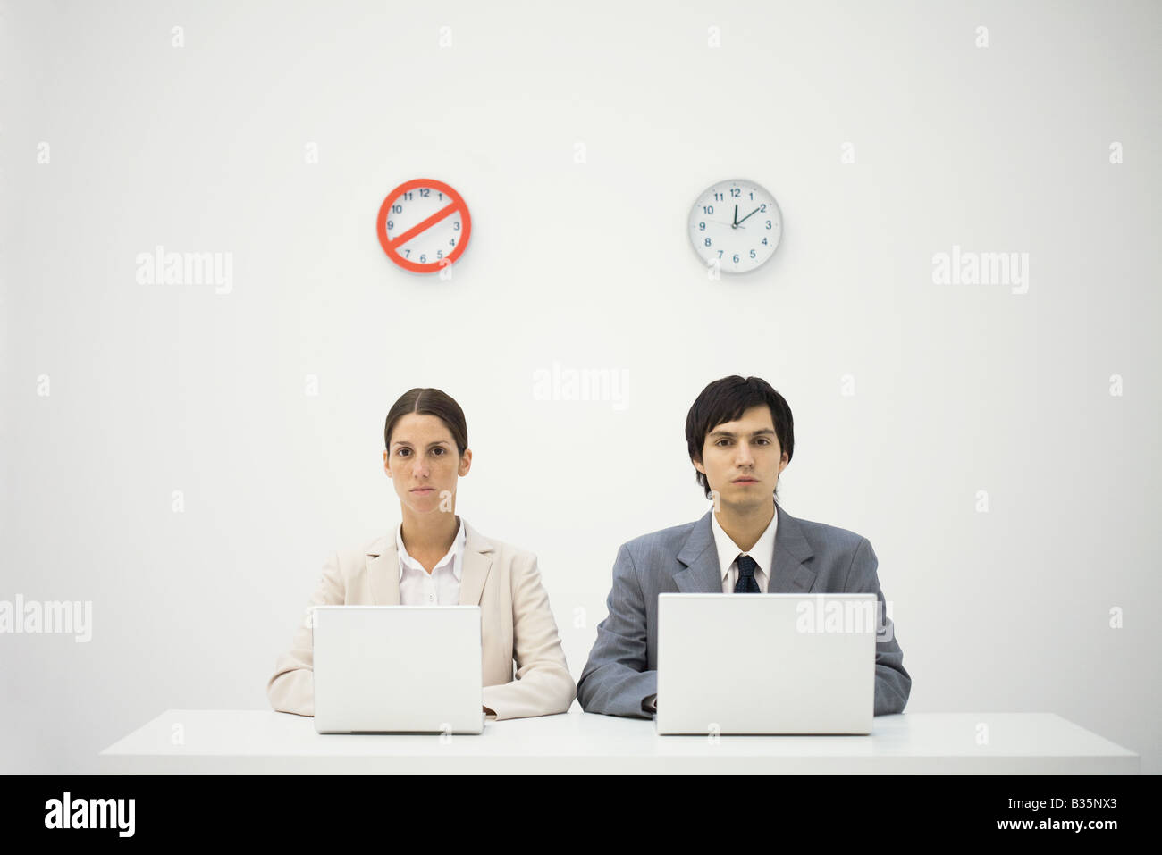 Office workers sitting below clocks, using laptops, warning sign over ...