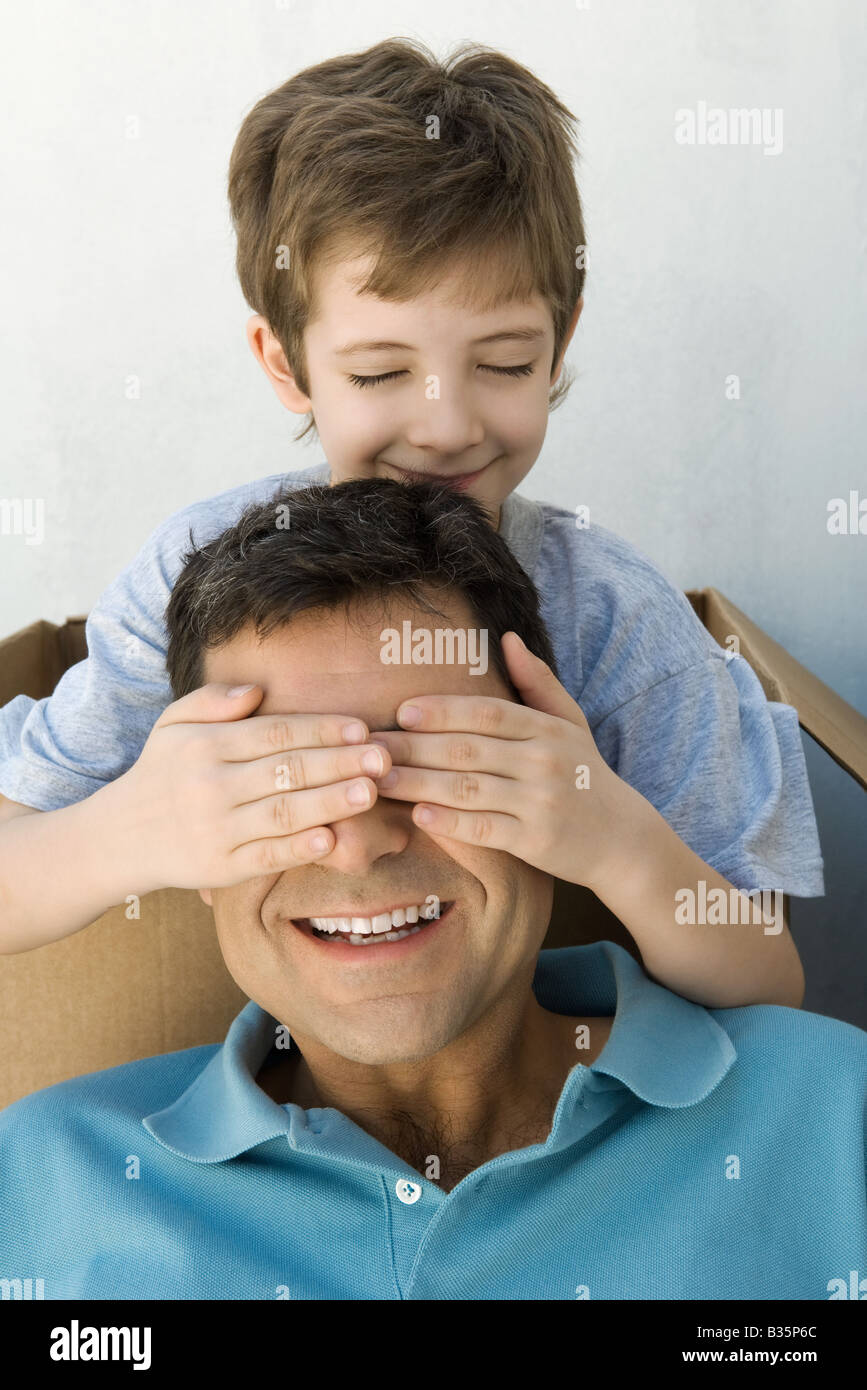 Boy with hands over his father's eyes, both smiling, boy's eyes closed ...