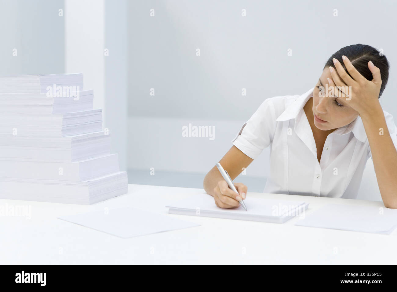 Woman holding her forehead, writing on blank sheet of paper, looking at ...