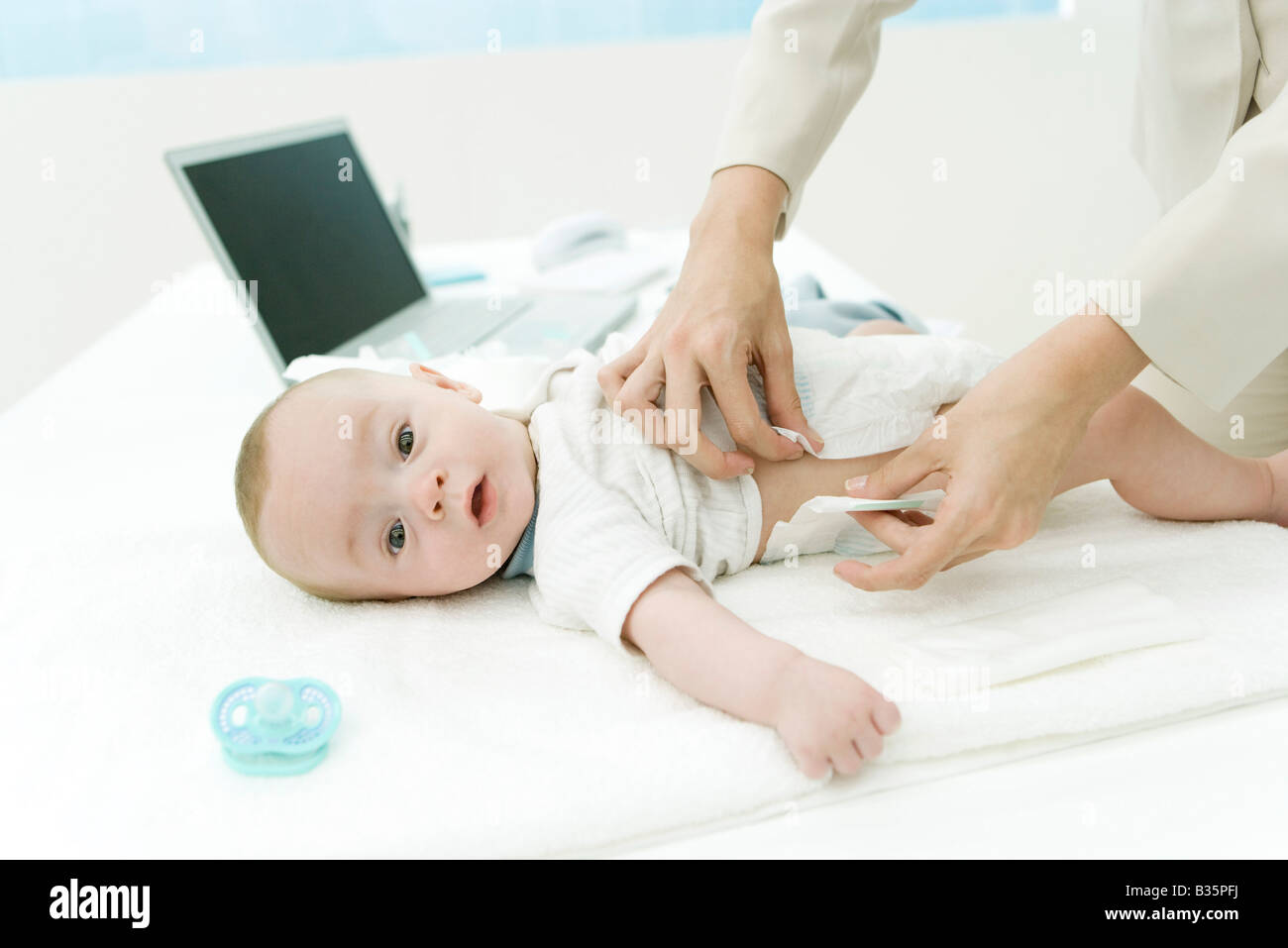 Mother changing baby's diaper on desk, laptop computer in background ...