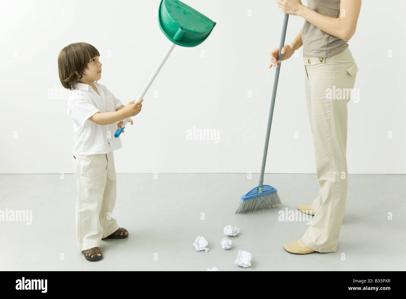 Little boy playing with dust pan while his mother is sweeping the floor ...