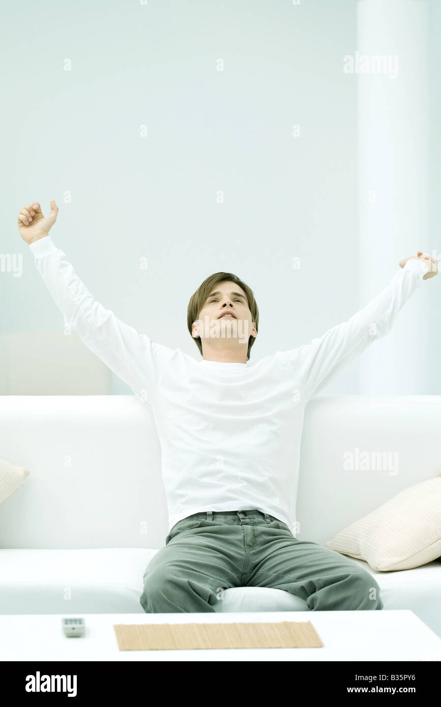 Young man sitting on couch with arms raised Stock Photo - Alamy