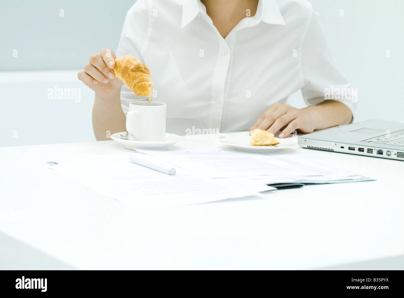 Woman dipping croissant into coffee, sitting at cluttered desk, cropped ...