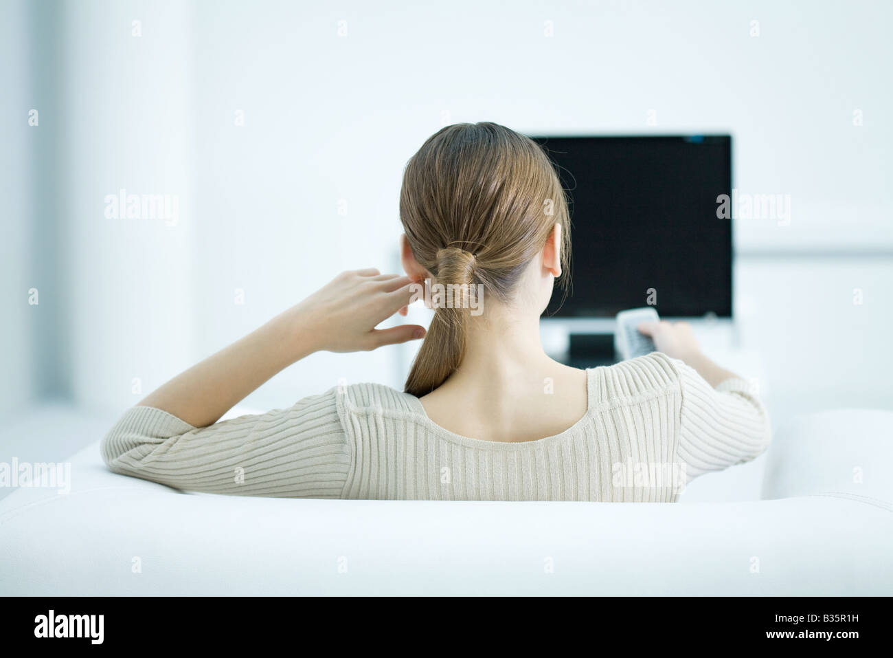 Young woman sitting in armchair watching TV, holding remote, rear view ...