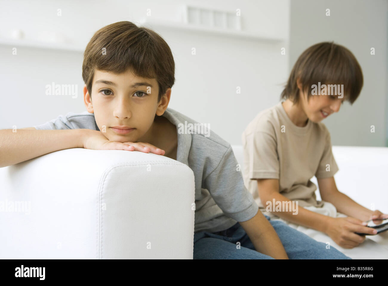 Boys sitting on couch, one looking at camera, the other playing ...