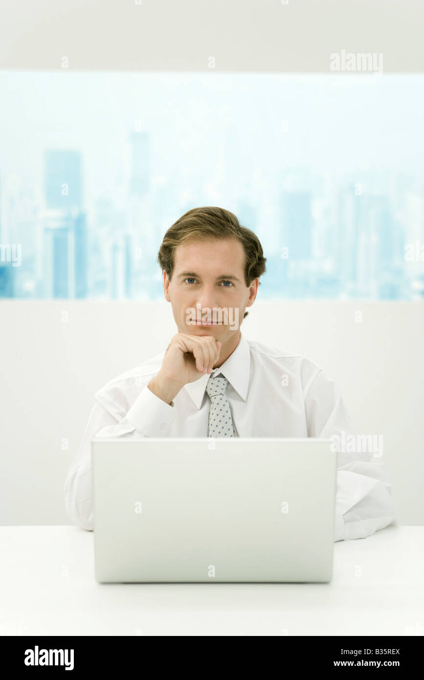 Businessman sitting with laptop computer, holding head, smiling at ...