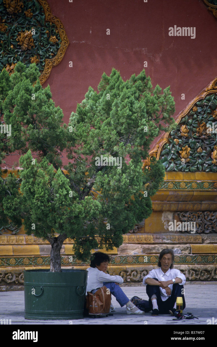 Two women sit in shade of containerised tree by wall in Forbidden City ...