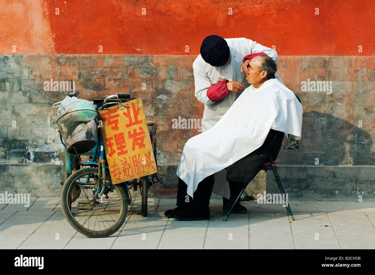 China, Beijing. An outdoor barber cutting hair beside a temple wall ...
