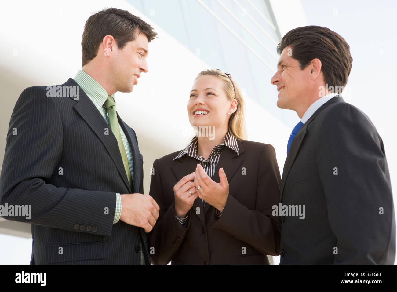 Three businesspeople standing outdoors by building talking and smiling ...
