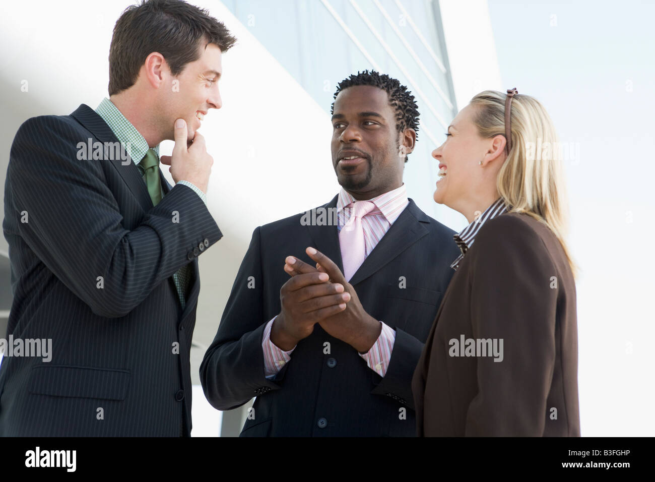 Three businesspeople standing outdoors by building talking and smiling ...