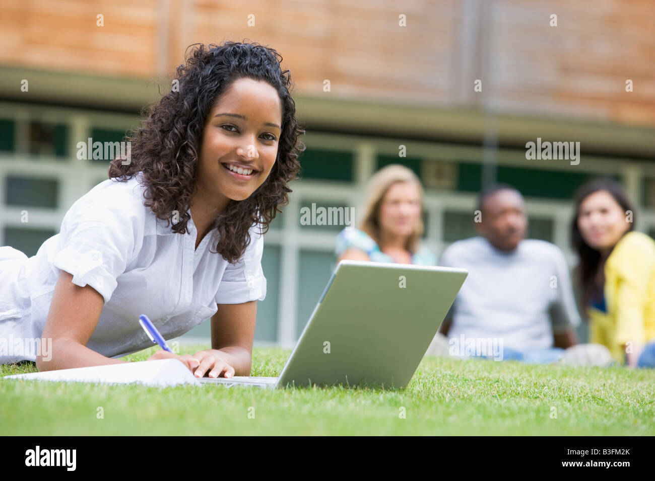 Student outdoors on lawn using laptop with other students in background ...