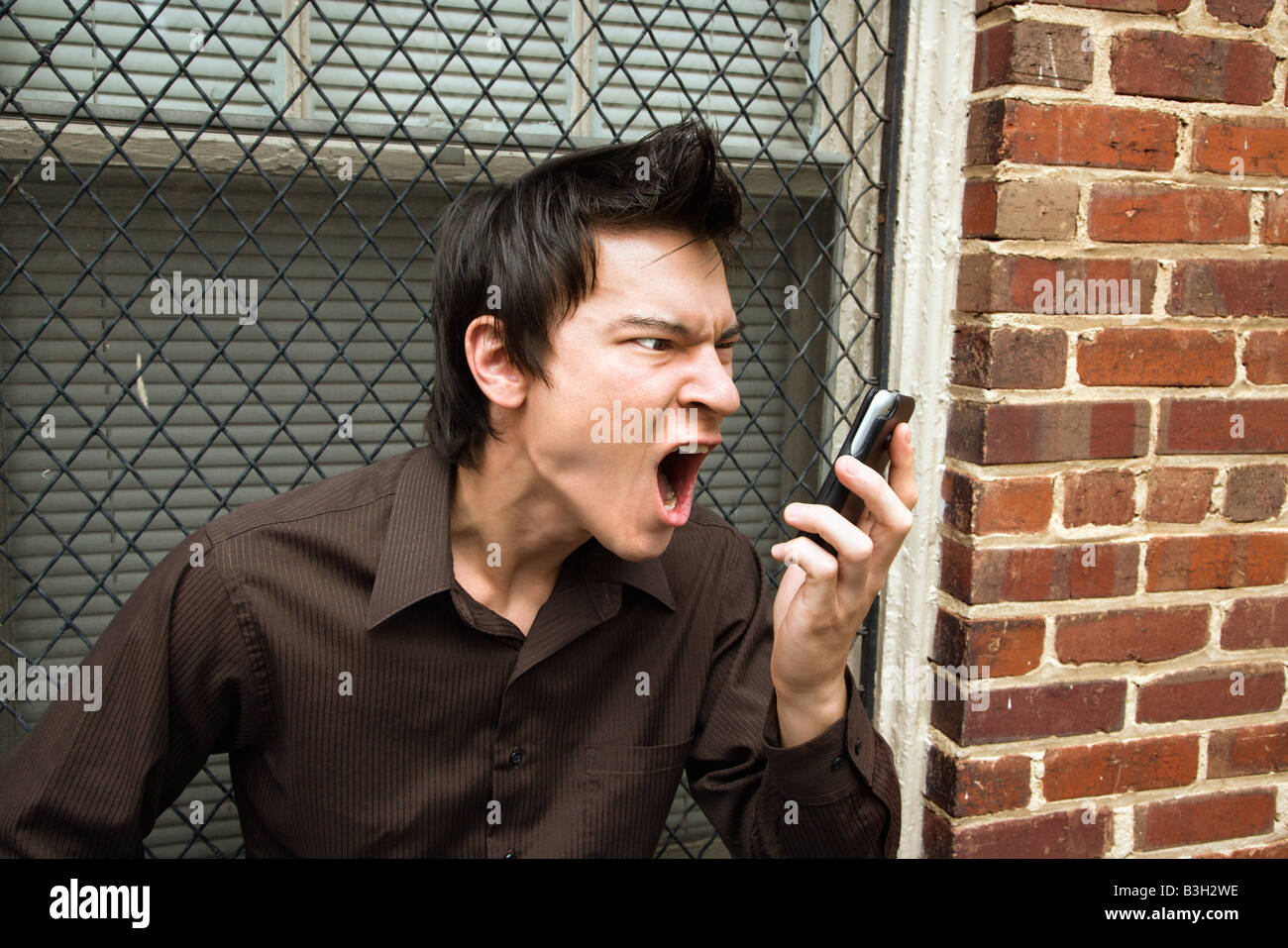 Angry young Asian man next to brick wall and window yelling at cell ...