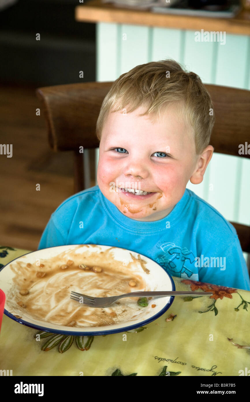 Young happy boy with food on his face and empty plate in front of him ...