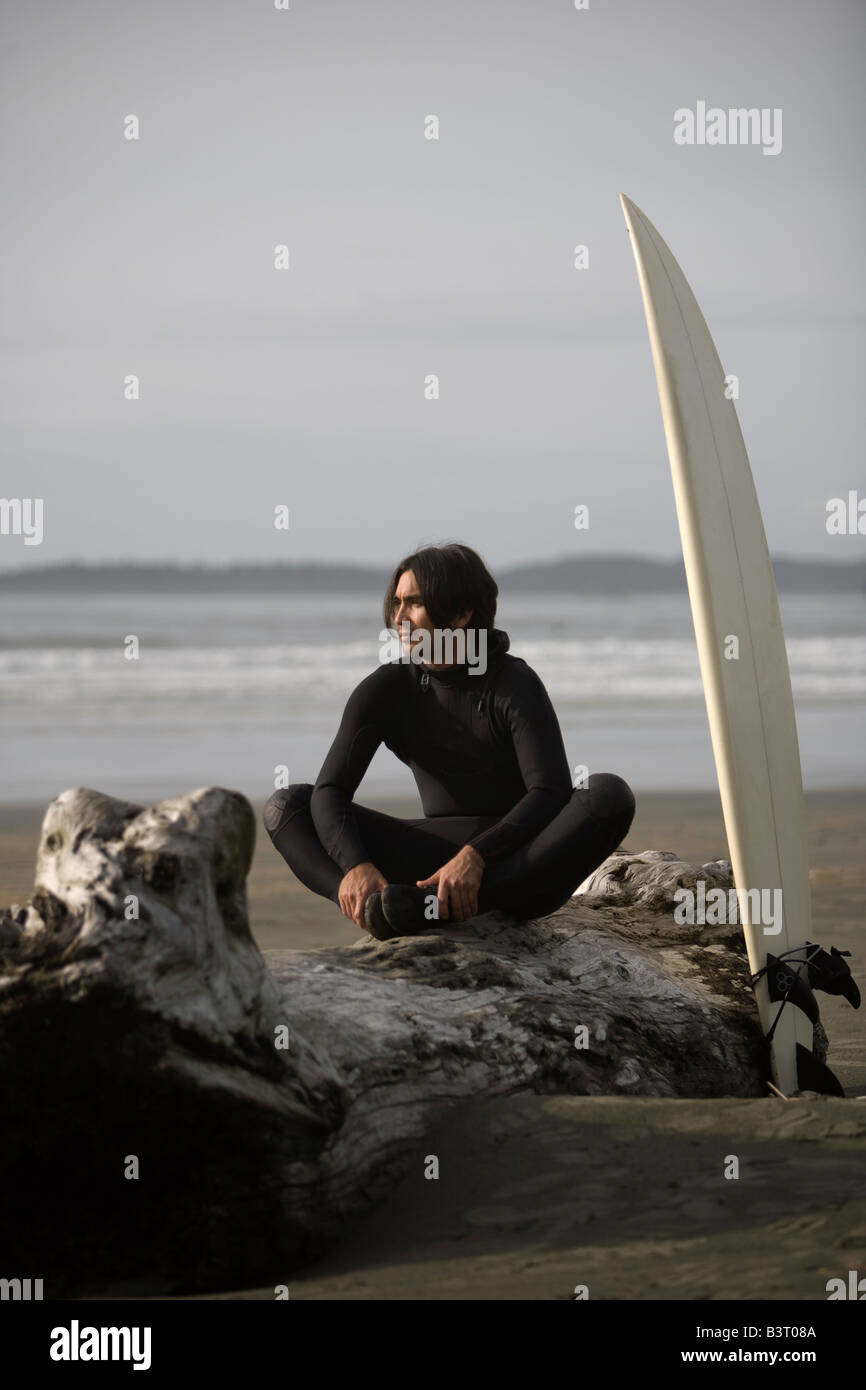Surfer sitting on log on beach, Cox Bay near Tofino, British Columbia ...