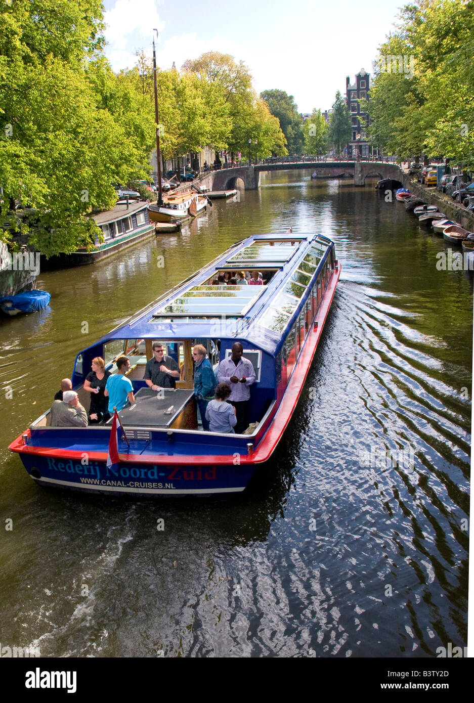 A tour boat full of people travel a canal lined with trees and house ...