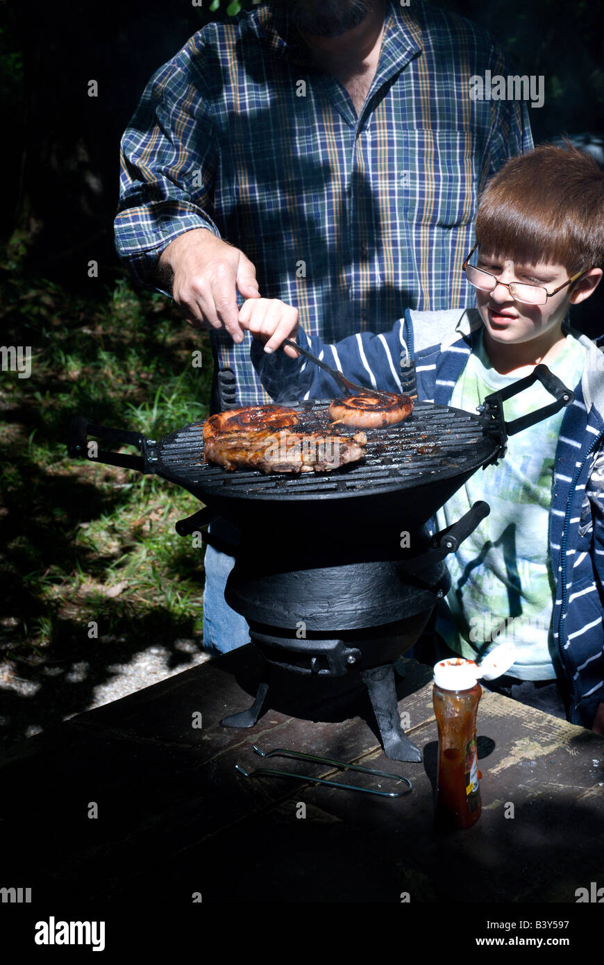 A boy is turning the meat on a barbeque grill with the helping hand of ...