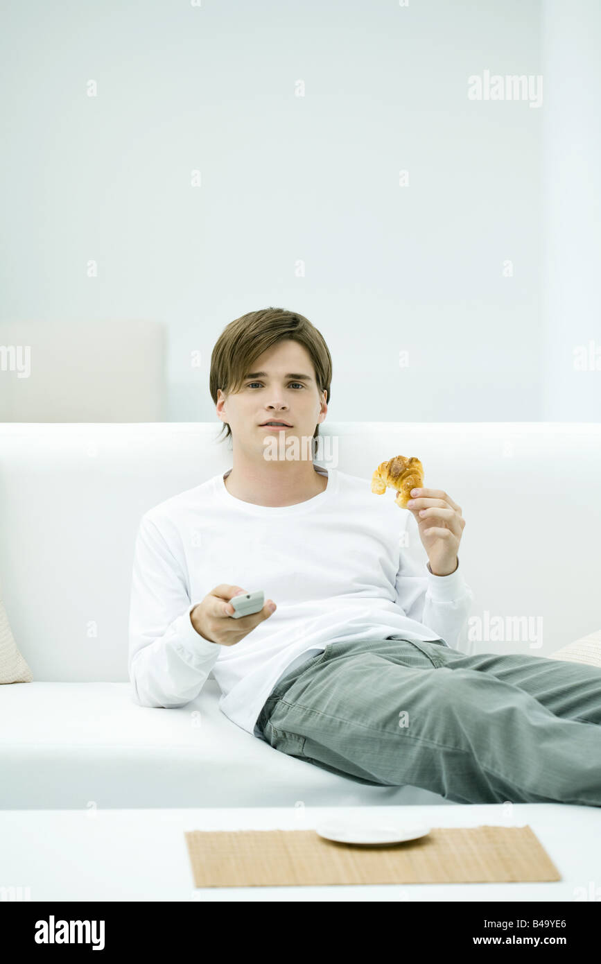 Young man sitting on sofa, holding remote control and croissant Stock ...