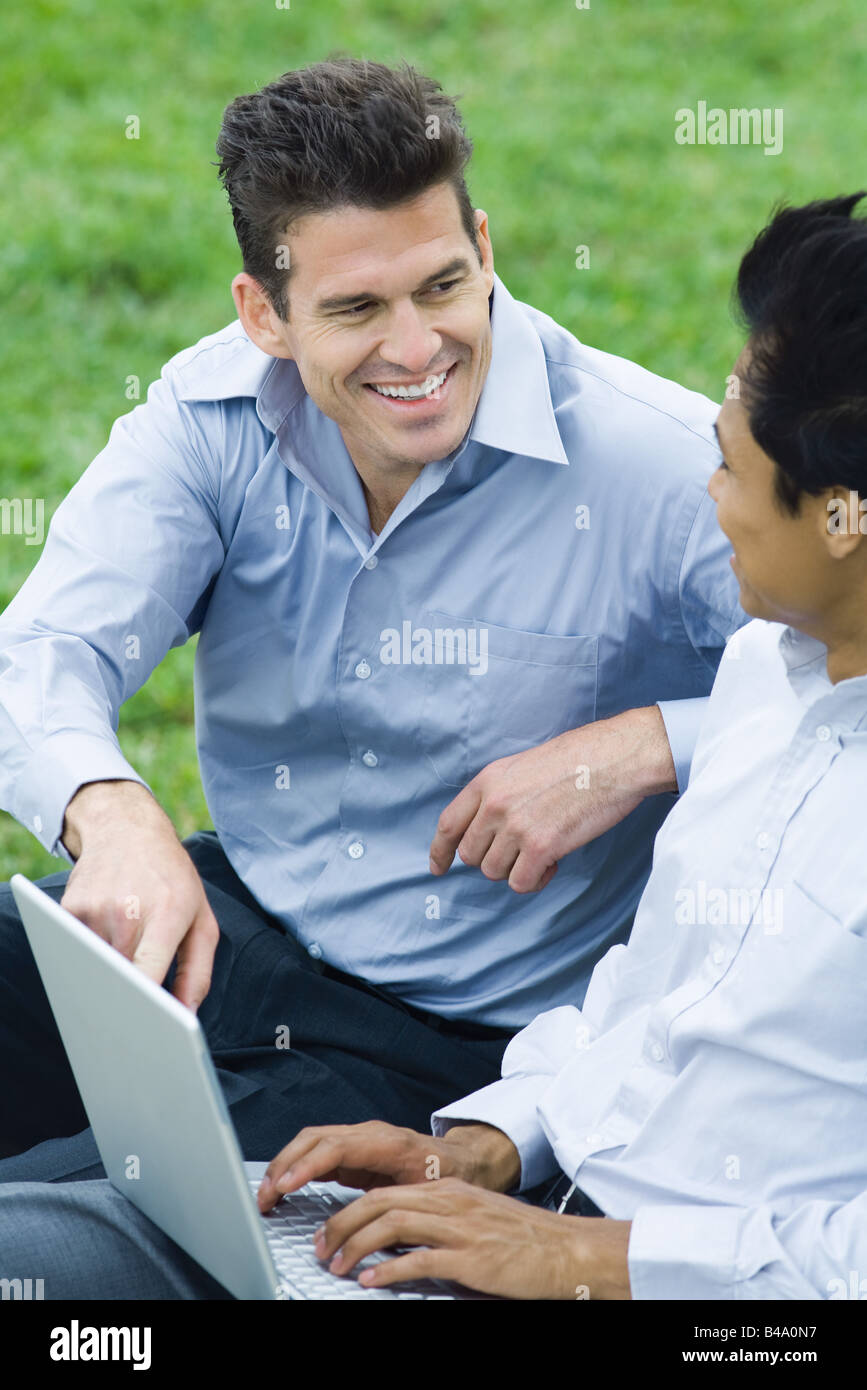 Two men sitting in park with laptop computer, looking at each other ...