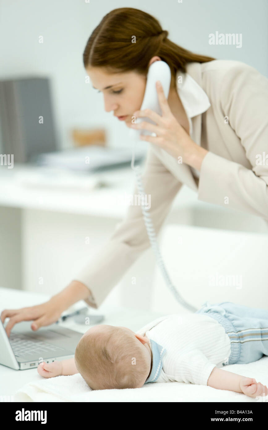 Professional woman using laptop computer and phone, infant lying on ...